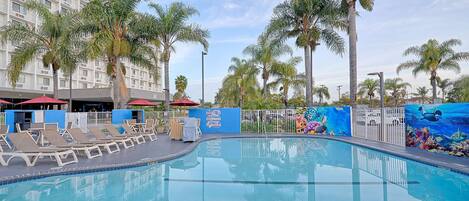 Piscina com espreguiçadeiras, palmeiras e um edifício de hotel ao fundo.