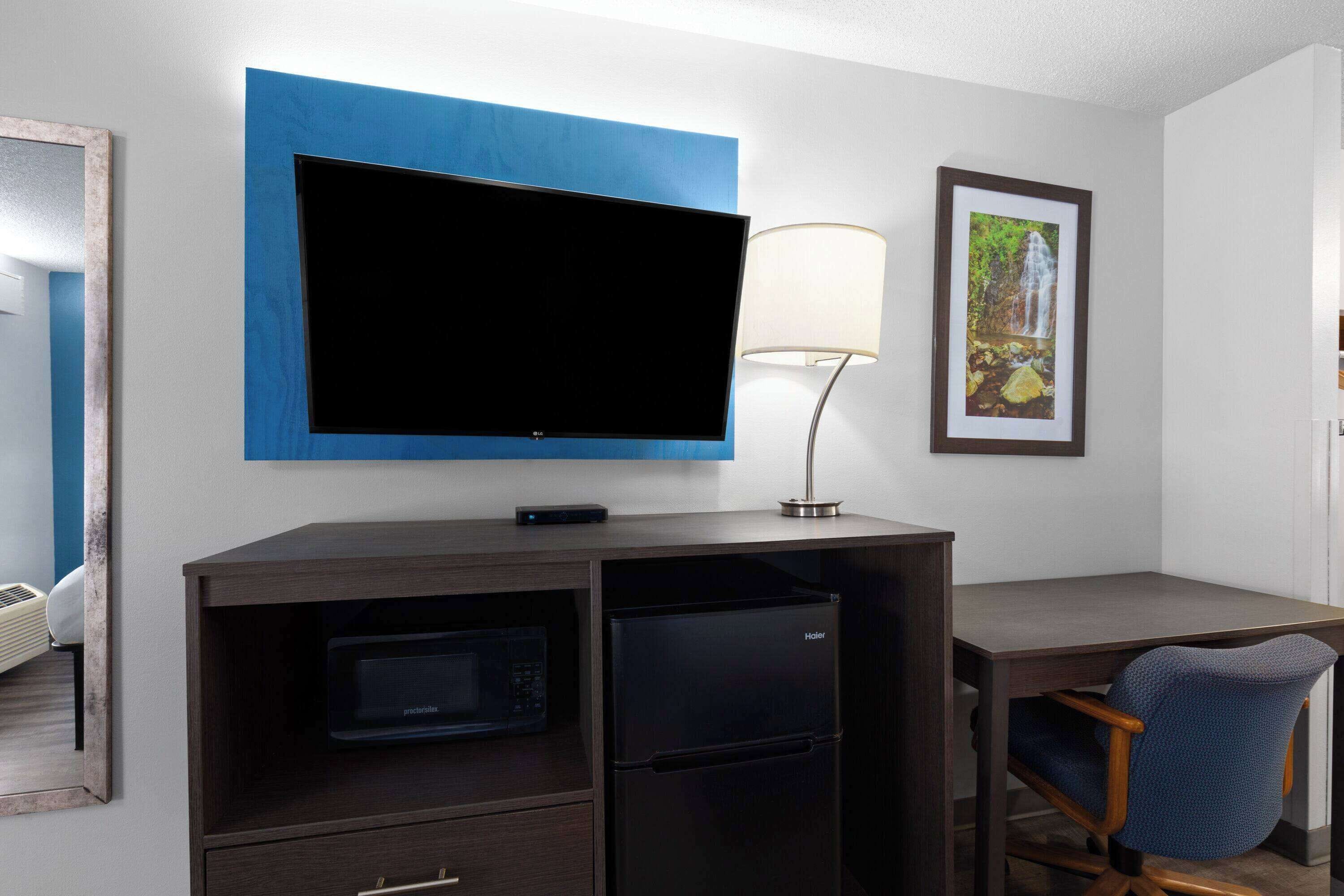 Quarto de hotel com televisão de tela plana na parede, uma escrivaninha com luminária, micro-ondas e geladeira.