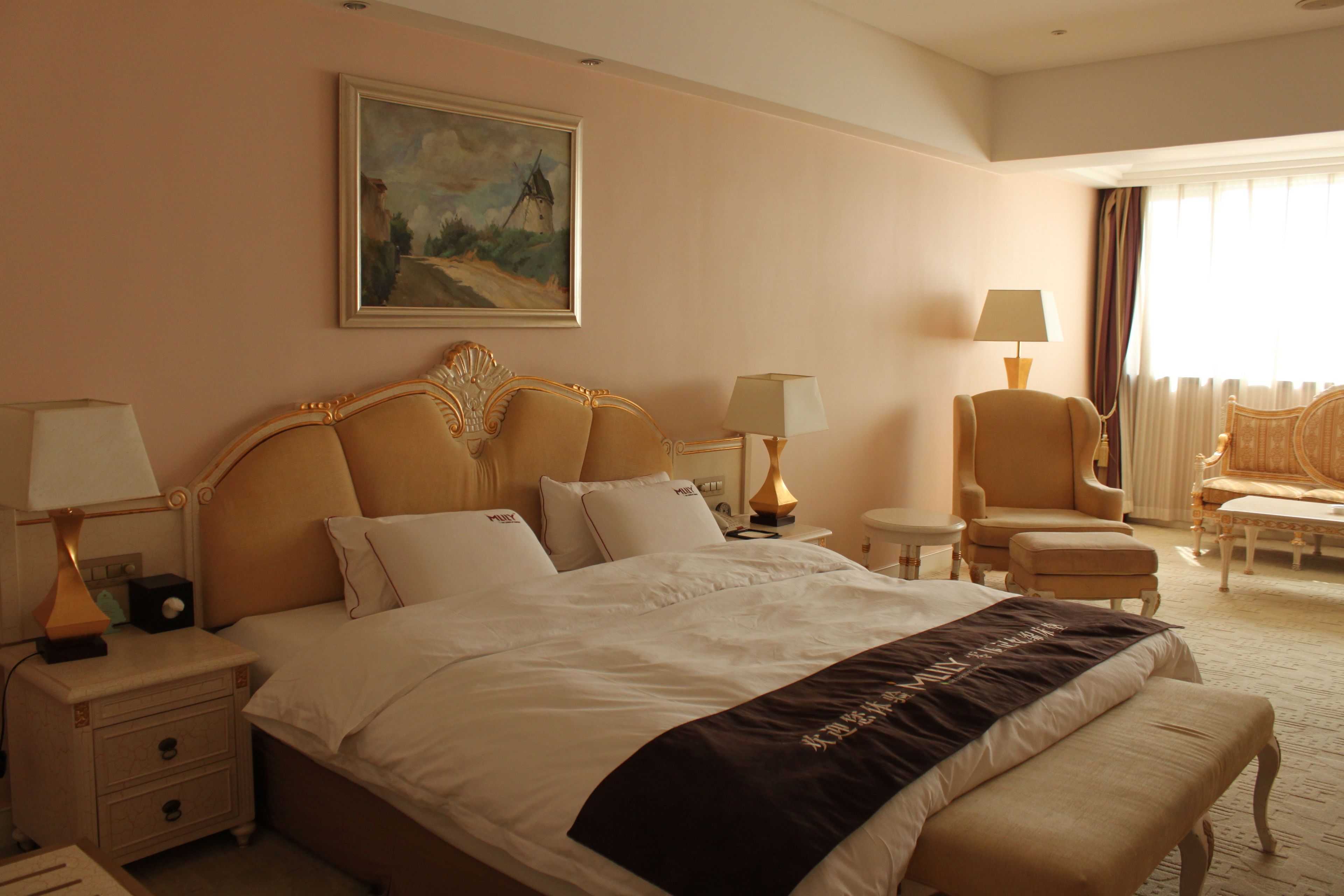 Quarto de hotel com uma cama grande, duas mesas de cabeceira com luminárias, uma cadeira, um banco e um quadro na parede.