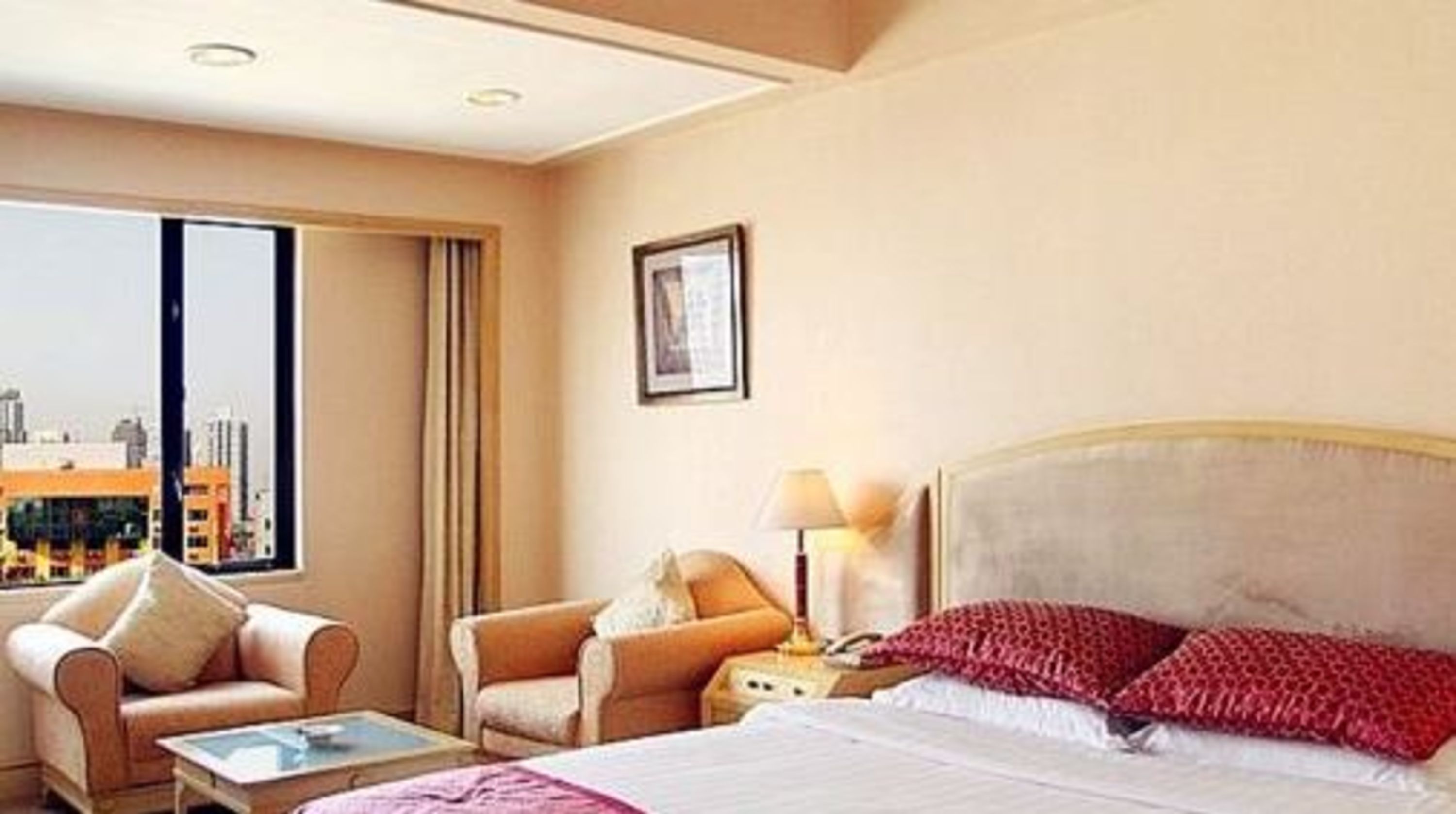 Quarto de hotel com cama, poltrona, mesa pequena, luminária e janela com vista para a cidade.