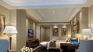 Quarto de hotel com cama, mesa de cabeceira, escrivaninha, cadeira, abajur e obras de arte nas paredes.