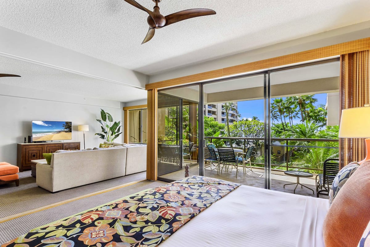 Quarto de hotel com cama, sofá, televisão, varanda com mesa e cadeiras, e vista para palmeiras.