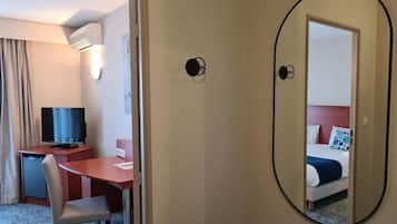 Quarto de hotel com cama, escrivaninha com televisão, cadeira, espelho e porta que dá para outro cômodo.