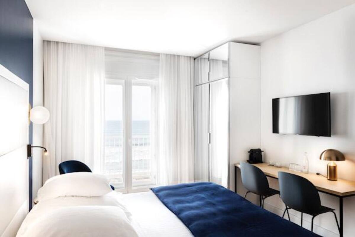 Quarto de hotel com cama, escrivaninha, duas cadeiras, televisão e vista da cidade através de amplas janelas.