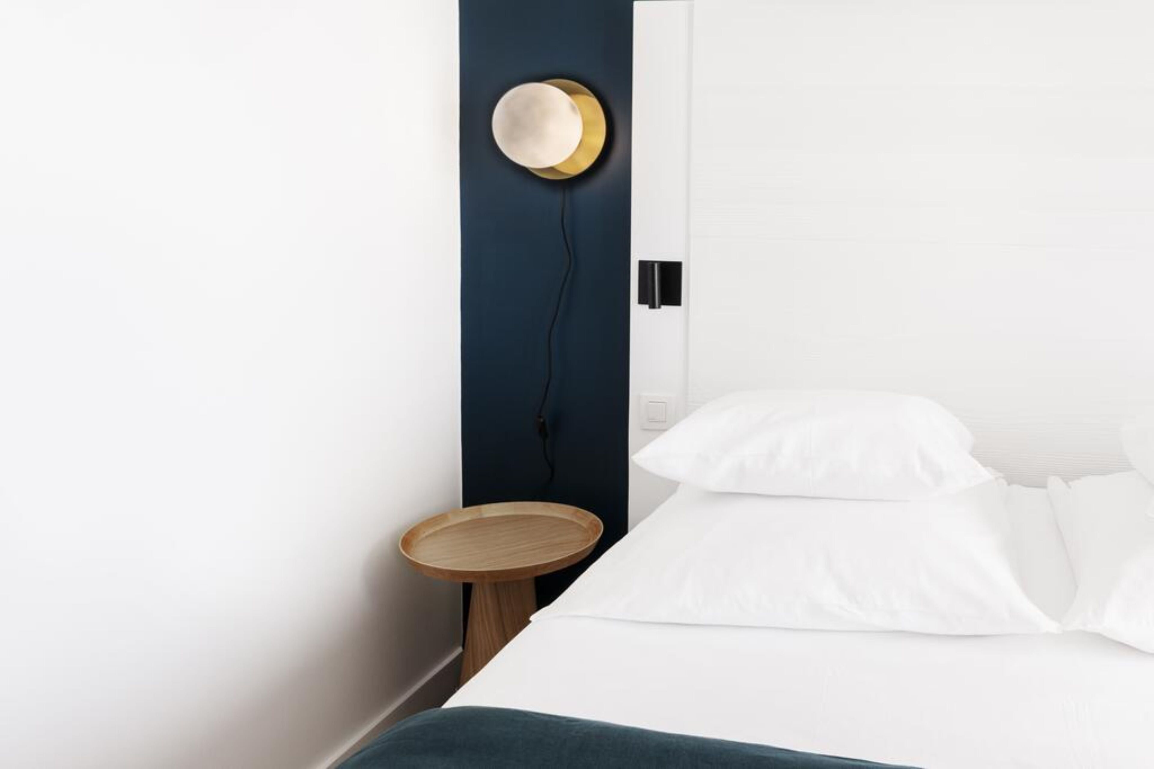 Quarto de hotel bem arrumado com cama, mesa lateral redonda de madeira, luminária fixada na parede e uma pequena tomada.