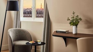 Quarto com vista para a Torre Eiffel, uma cadeira, uma pequena mesa com um copo de água e um livro, um abajur e uma planta em um prateleira.