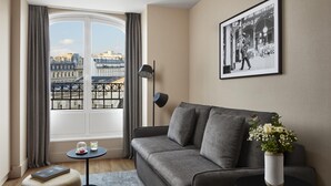 Uma sala de estar moderna com um sofá cinzento, uma mesa redonda pequena, um vaso com flores, uma fotografia a preto e branco emoldurada e vista para edifícios parisienses através de uma grande janela.
