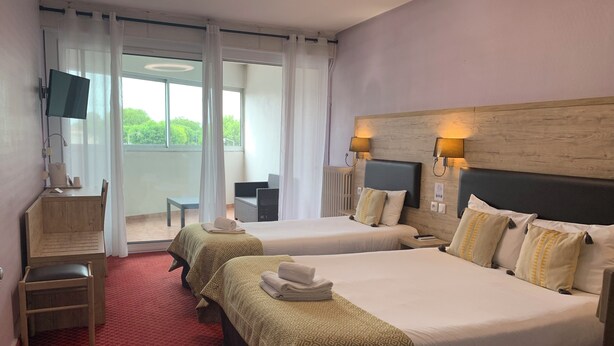 Quarto de hotel com duas camas, uma escrivaninha, uma cadeira, televisão e vista para o verde lá fora.
