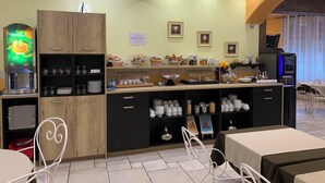 Espaço de café da manhã de hotel, com máquina de café, buffet de pães e doces, e área de estar com mesas e cadeiras.