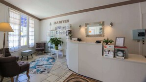 Uma área de recepção de hotel com balcão, quadros decorativos, planta em vaso e um espaço de estar com cadeiras e mesa.