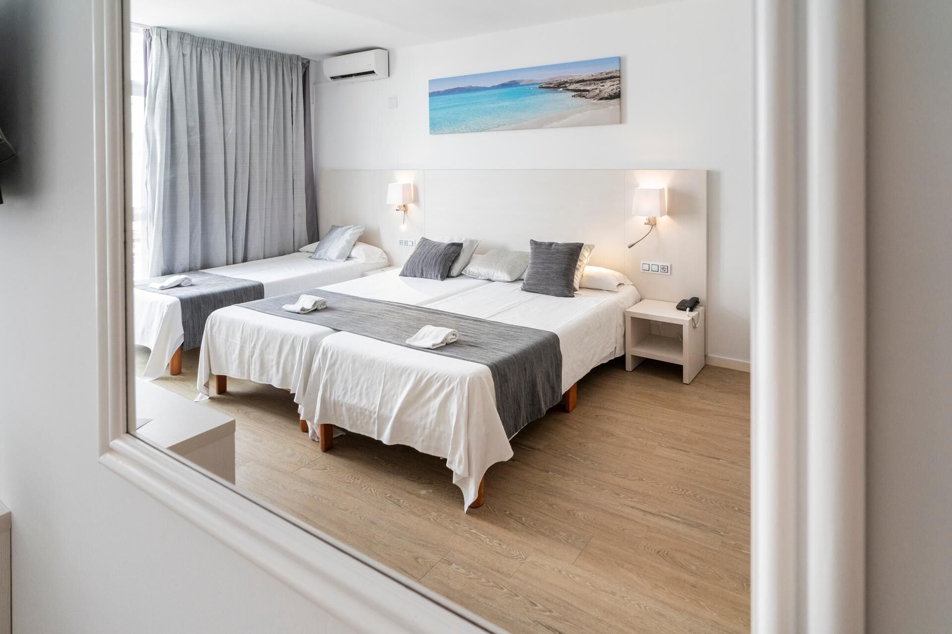 Quarto de hotel com duas camas, uma janela grande com cortinas, um quadro de uma praia e uma mesa de cabeceira com abajur.