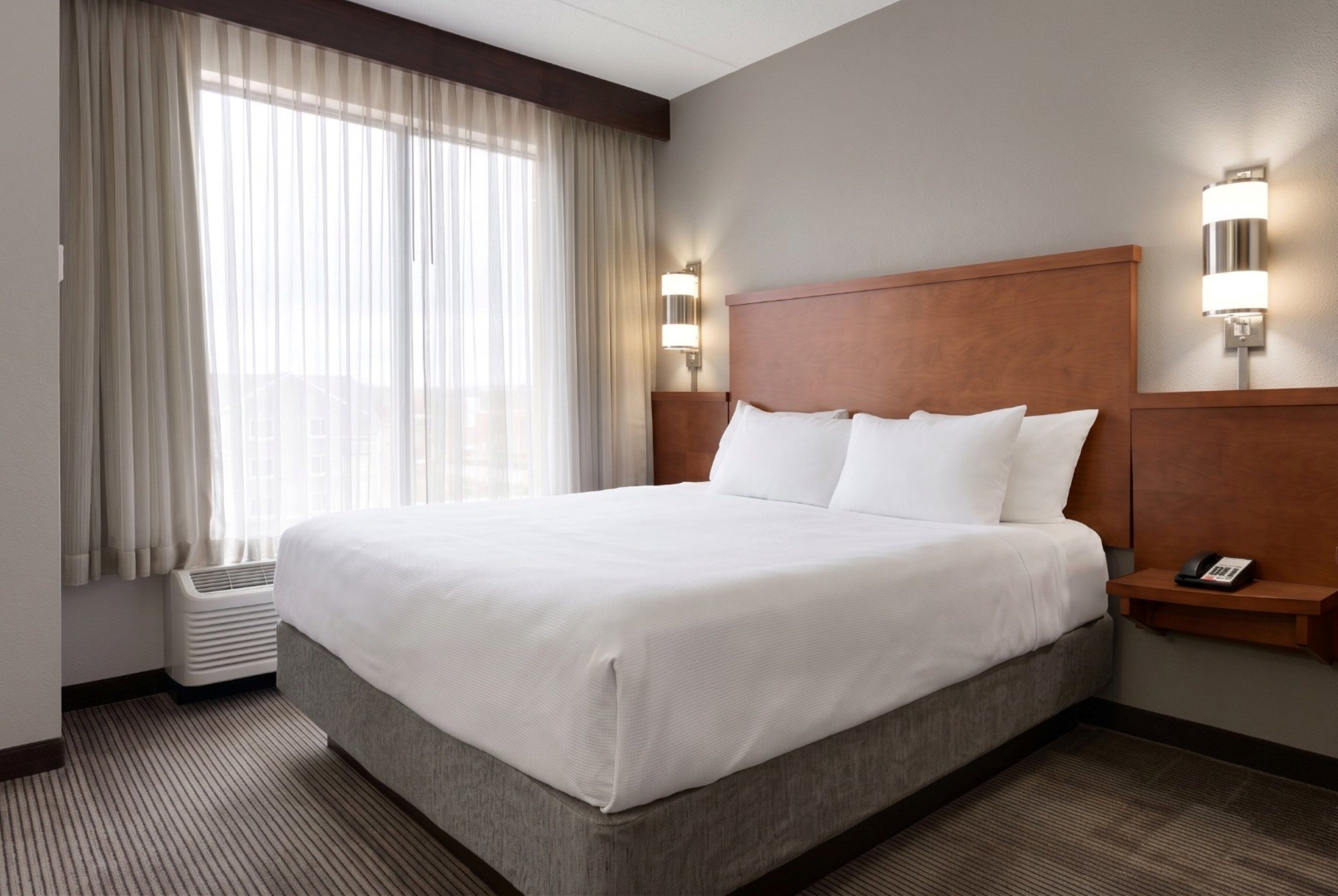Quarto de hotel com uma cama grande, uma mesa de cabeceira com telefone, uma luminária na parede e uma janela com cortinas leves.