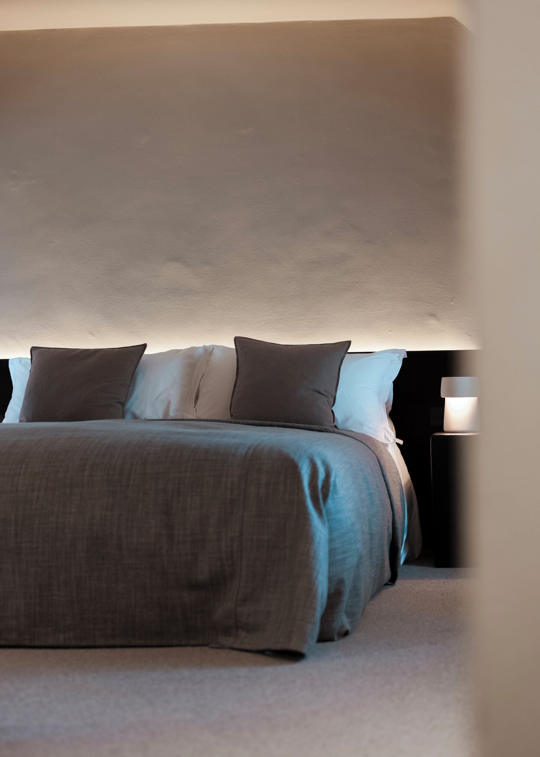 Quarto moderno com uma cama grande, várias almofadas e uma mesa de cabeceira com abajur.