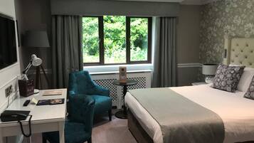 Quarto de hotel com cama, escrivaninha, cadeira, janela com cortinas, luminária e vista para o verde lá fora.