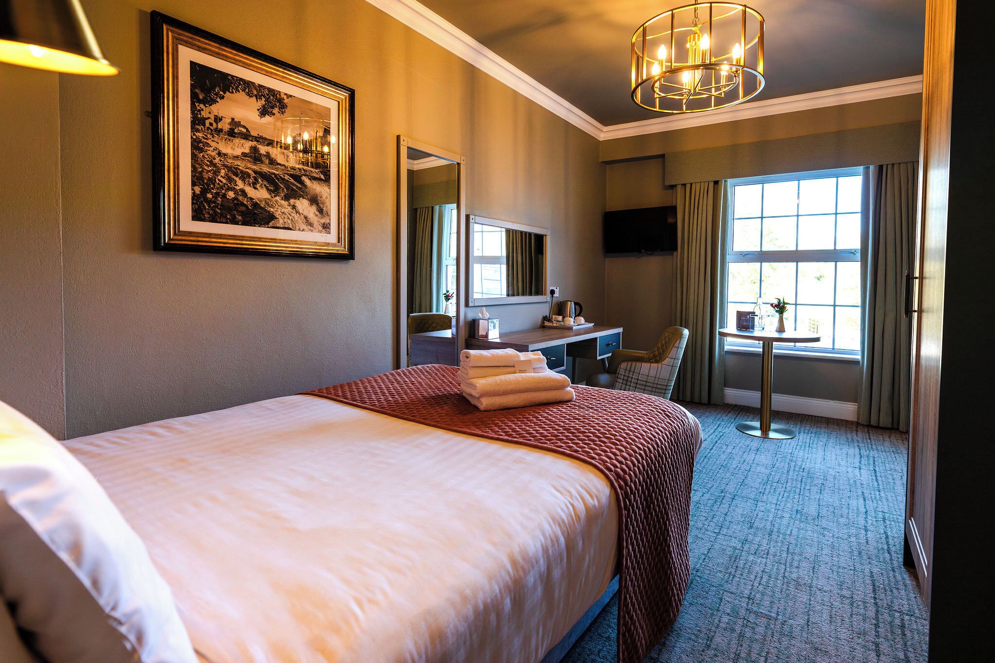 Quarto de hotel com cama, escrivaninha, cadeira, televisão e janela.