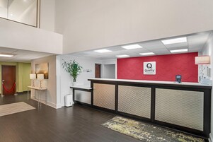 Uma área de recepção de hotel com parede vermelha, balcão de atendimento, planta e tapete vermelho.