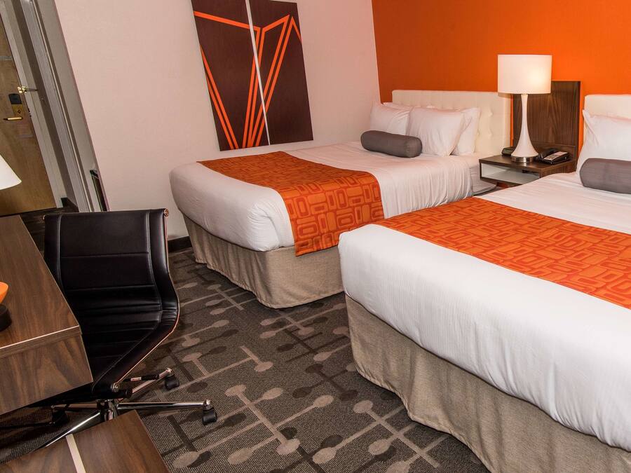 Quarto de hotel com duas camas, uma escrivaninha, uma cadeira, um abajur e uma parede com arte geométrica.