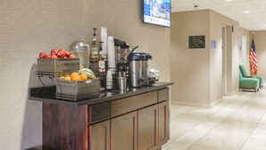 Uma área de recepção de hotel com um balcão de buffet repleto de frutas, bebidas e máquinas de café.