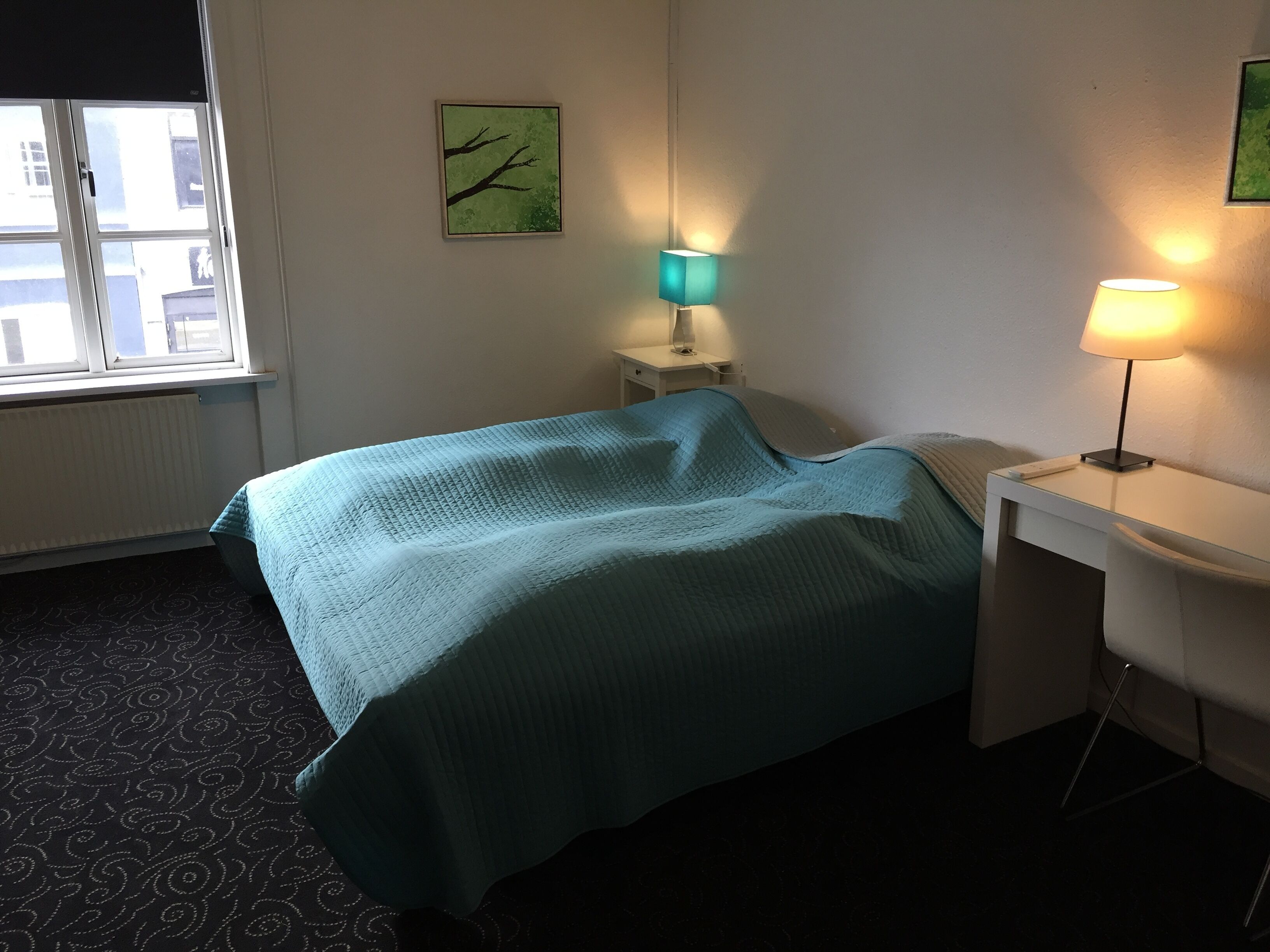 Quarto com cama, escrivaninha, luminária, janela e uma obra de arte na parede.