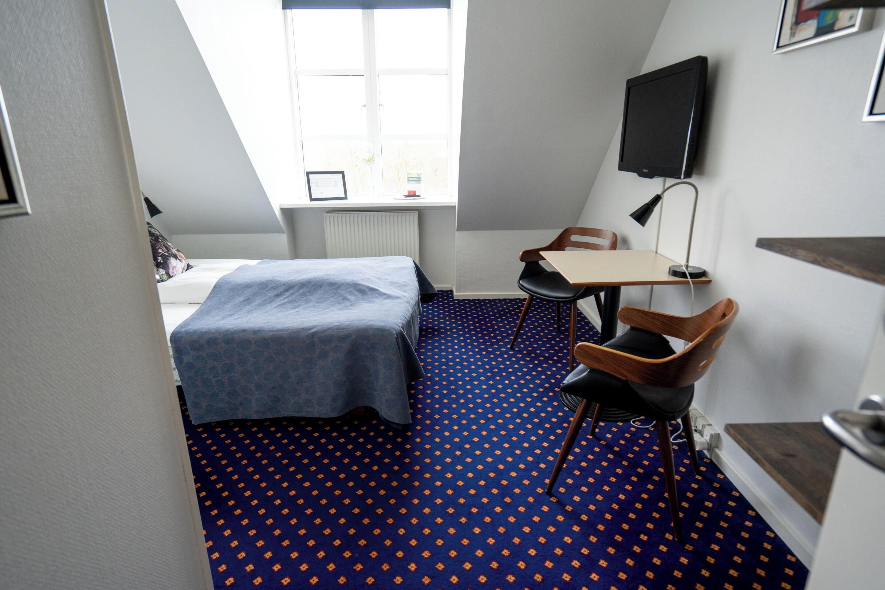 Quarto pequeno e compacto com cama, escrivaninha, cadeira e televisão.