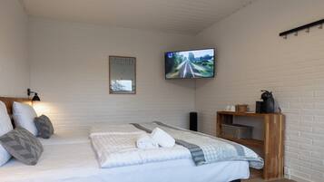Quarto de hotel com duas camas, uma televisão fixada na parede, uma prateleira de madeira com uma chaleira e uma cesta.