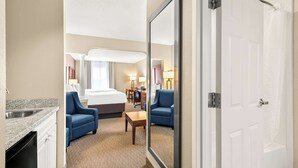 Quarto de hotel com cama, duas poltronas, uma mesinha, espelho, porta e cozinha compacta com pia e fogão.