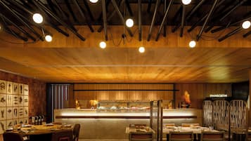 Interior moderno de restaurante com vigas de madeira no teto, área de bar central e disposição de mesas para os hóspedes.
