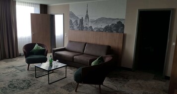 Quarto de hotel com um sofá castanho, duas poltronas, uma mesa de centro e uma pintura de uma igreja na parede.