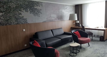 Quarto de hotel moderno com sofá de couro preto, poltrona vermelha e preta, mesa redonda pequena e escrivaninha com luminária.