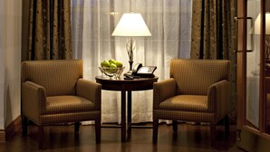 Quarto de hotel com duas poltronas, uma mesinha com abajur e vista através de cortinas leves.