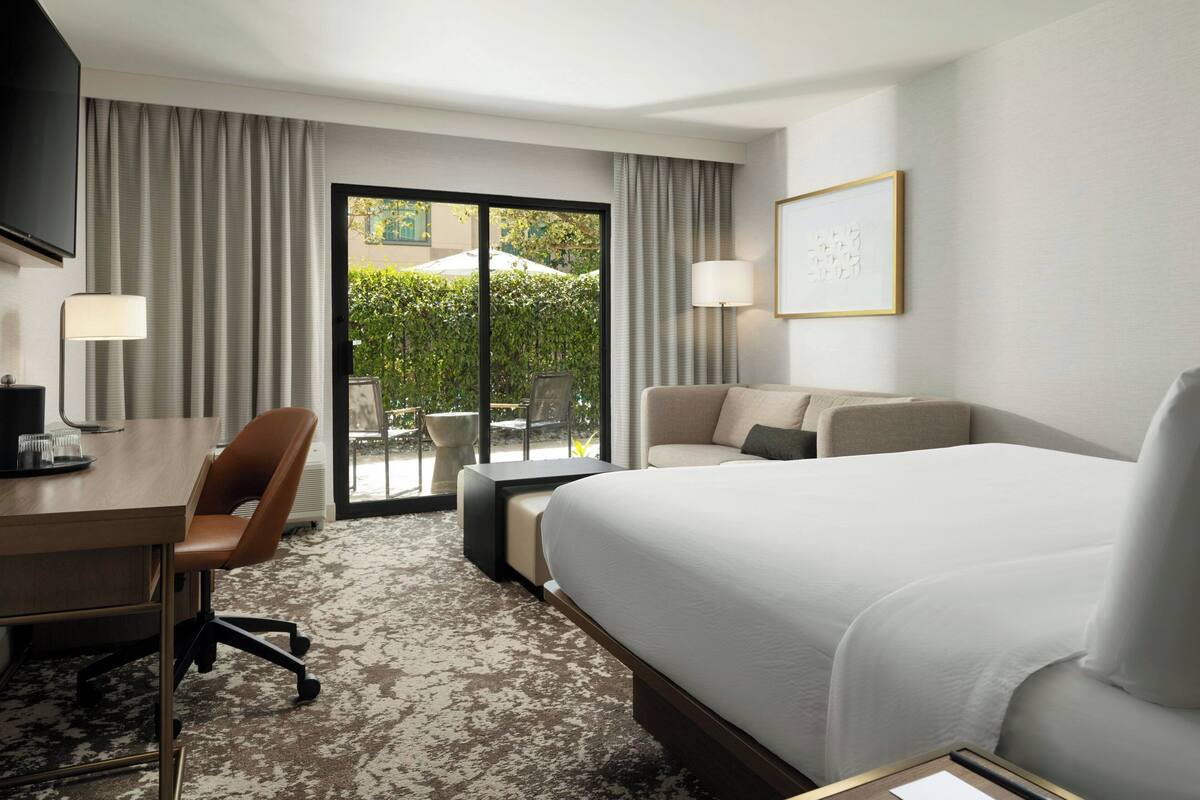 Quarto de hotel com cama, escrivaninha, cadeira, sofá e vista para o verde pela janela.