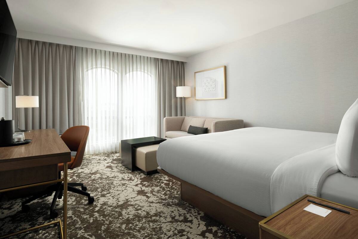 Quarto de hotel com uma cama grande, uma escrivaninha, uma cadeira, uma mesa pequena e uma luminária.