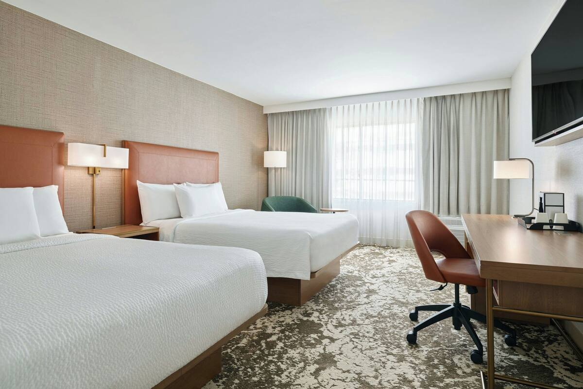 Quarto de hotel com duas camas, uma escrivaninha, uma cadeira e uma televisão.