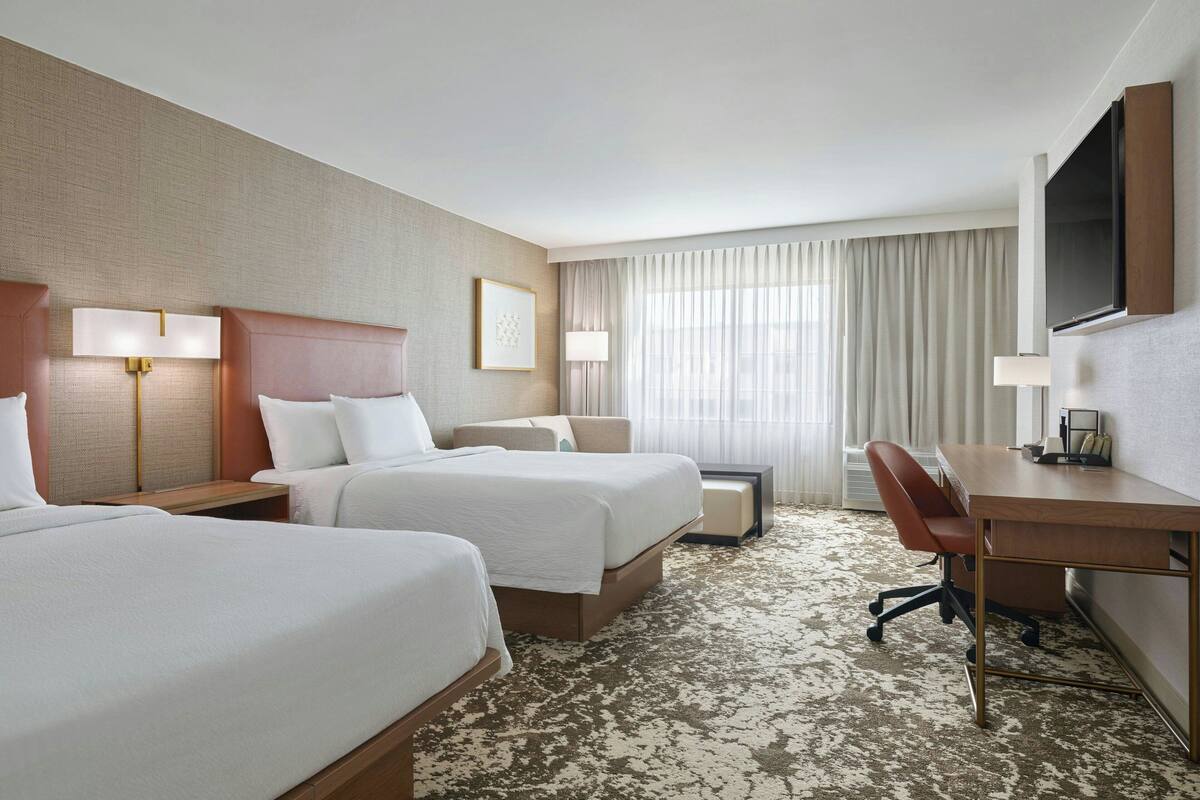 Quarto de hotel com duas camas, uma escrivaninha, uma cadeira, televisão e uma janela grande com cortinas.