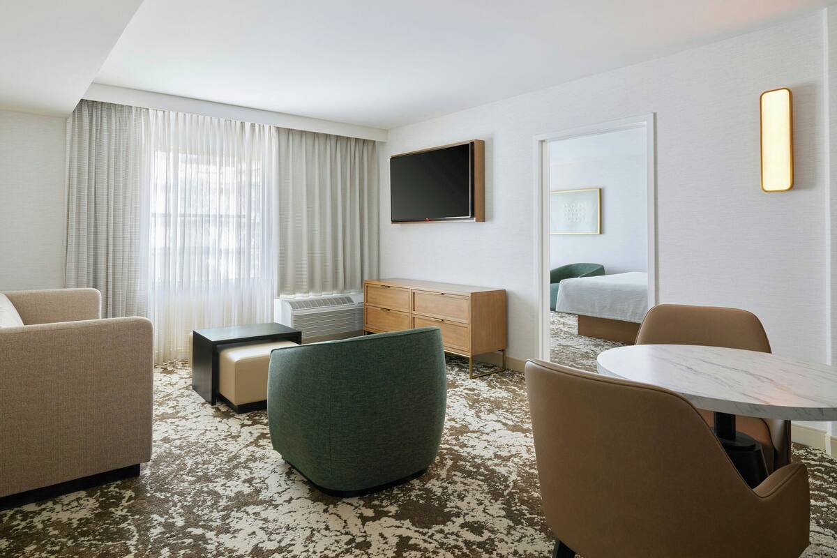 Quarto de hotel com uma cama grande, uma escrivaninha, uma cadeira, um sofá e uma televisão presa à parede.