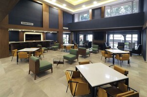 Um espaço amplo e interno, com várias mesas e cadeiras, uma tela grande e vista para o exterior através de grandes janelas.