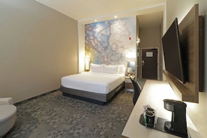 Quarto de hotel com cama, mesas de cabeceira, escrivaninha com cafeteira, televisão e parede com mural decorativo.