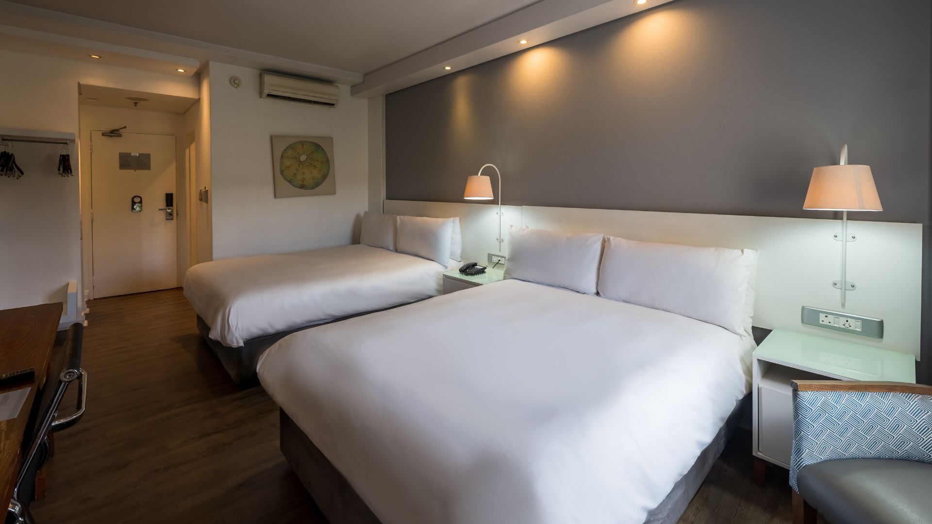 Quarto de hotel com duas camas, uma escrivaninha, uma cadeira, um abajur e um ar condicionado fixado na parede.
