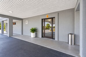 Entrada de prédio com teto plano, vaso de planta, lixeira e porta com uma placa em cima.