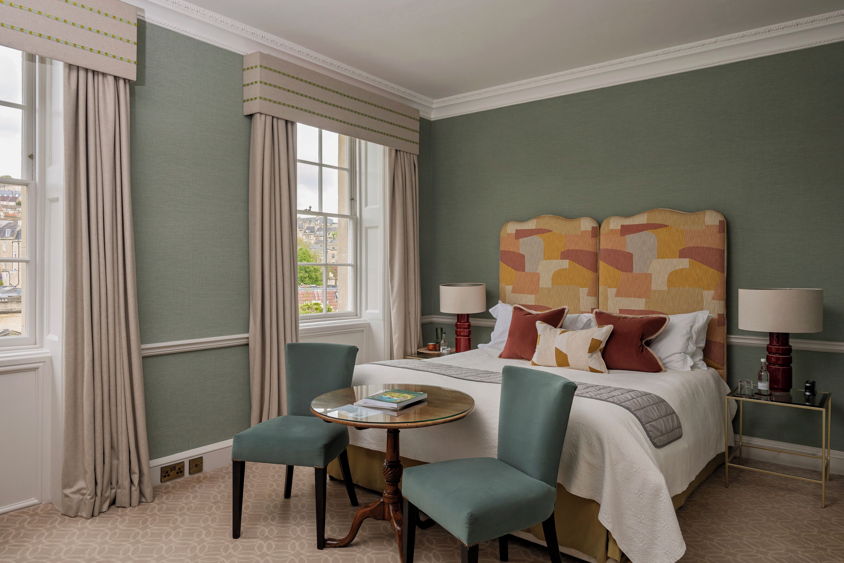 Quarto de hotel com cama, duas cadeiras, uma mesinha, mesa de cabeceira e janela com cortinas.