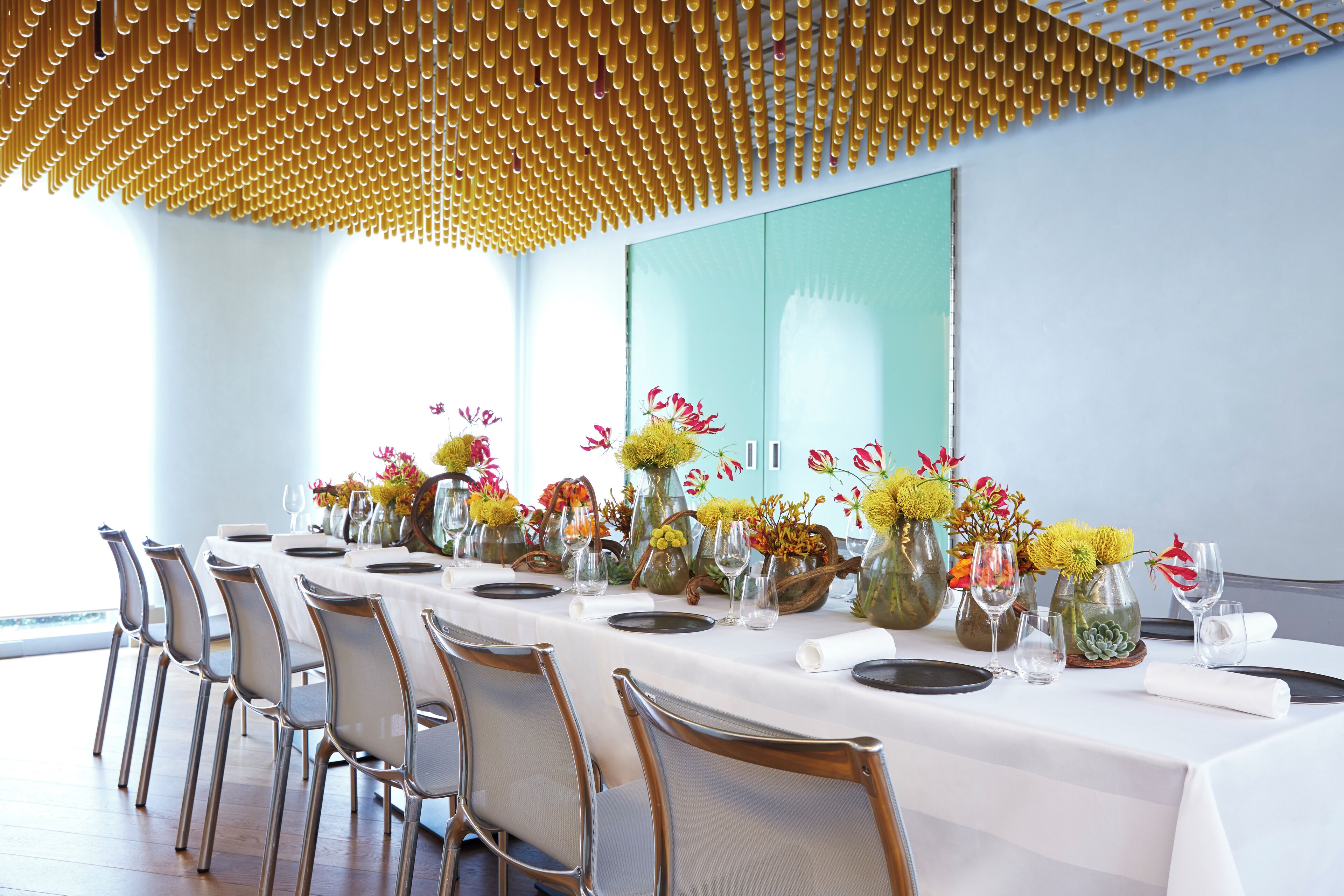 Uma longa mesa de jantar posta com cadeiras, vasos com flores e um design moderno no teto.
