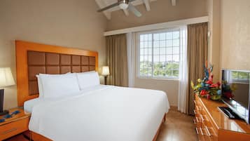 Quarto de hotel com uma cama grande, um ventilador de teto, uma televisão, uma escrivaninha com luminária e uma janela com cortinas.