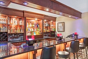 Uma área de bar com várias prateleiras de garrafas de bebidas, um balcão comprido e banquetas.