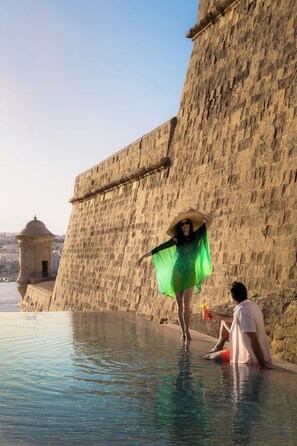 Área da piscina com parede de pedra e um pequeno edifício ao fundo.