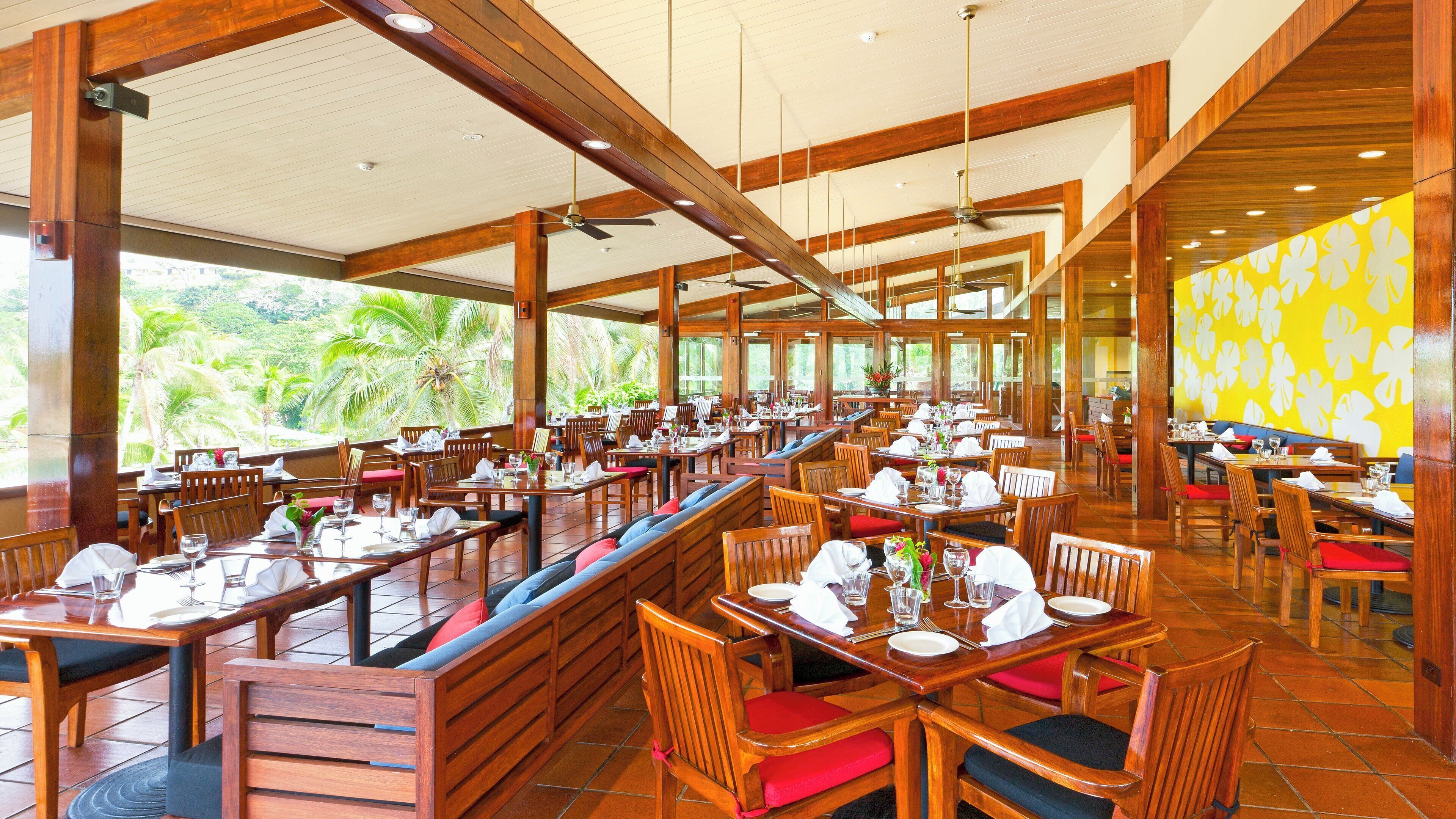 Um restaurante com móveis de madeira, grandes janelas e vista para palmeiras.