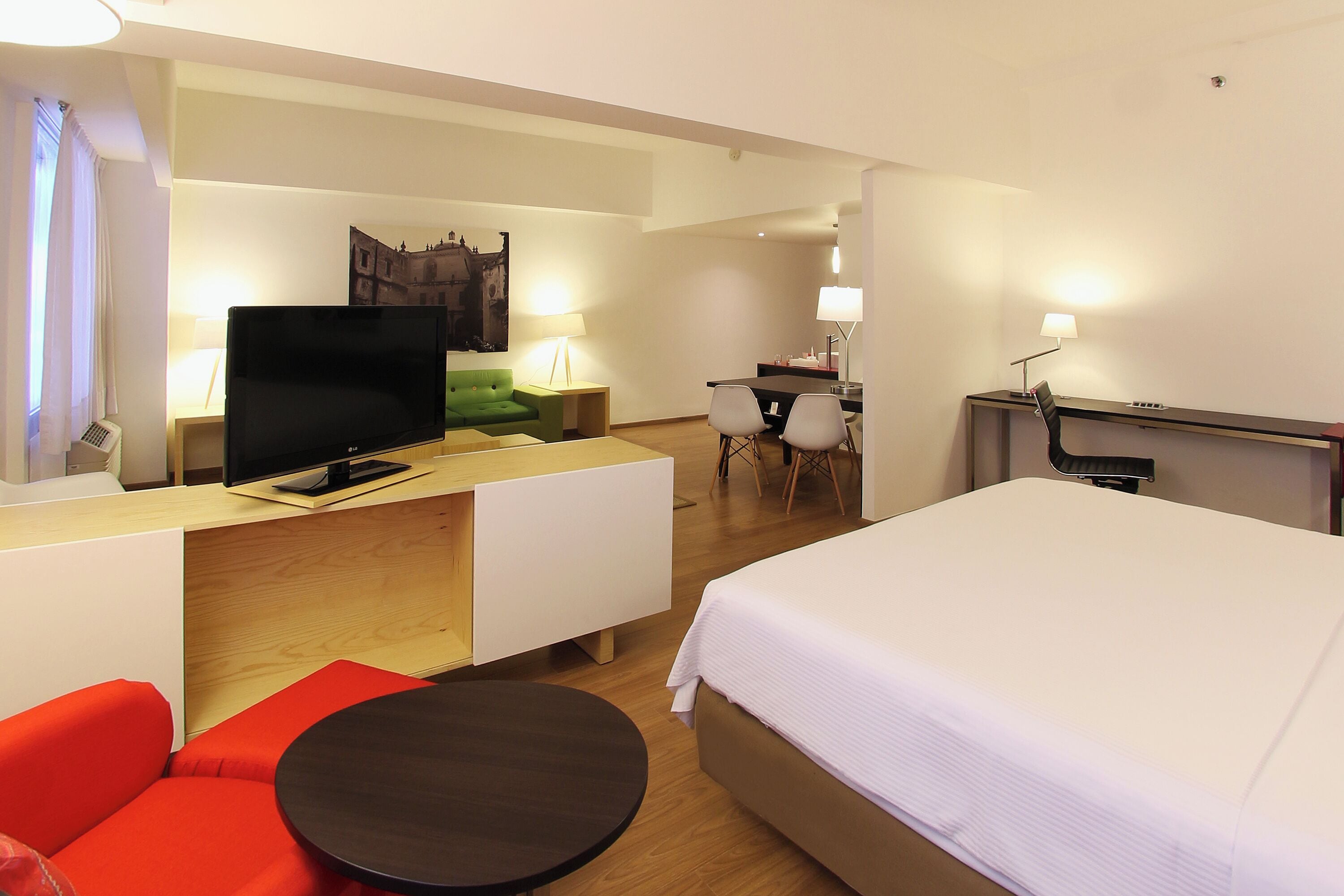 Quarto de hotel com cama, escrivaninha com computador, televisão e uma cadeira vermelha.