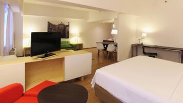 Quarto de hotel com cama, escrivaninha com computador, televisão e uma cadeira vermelha.