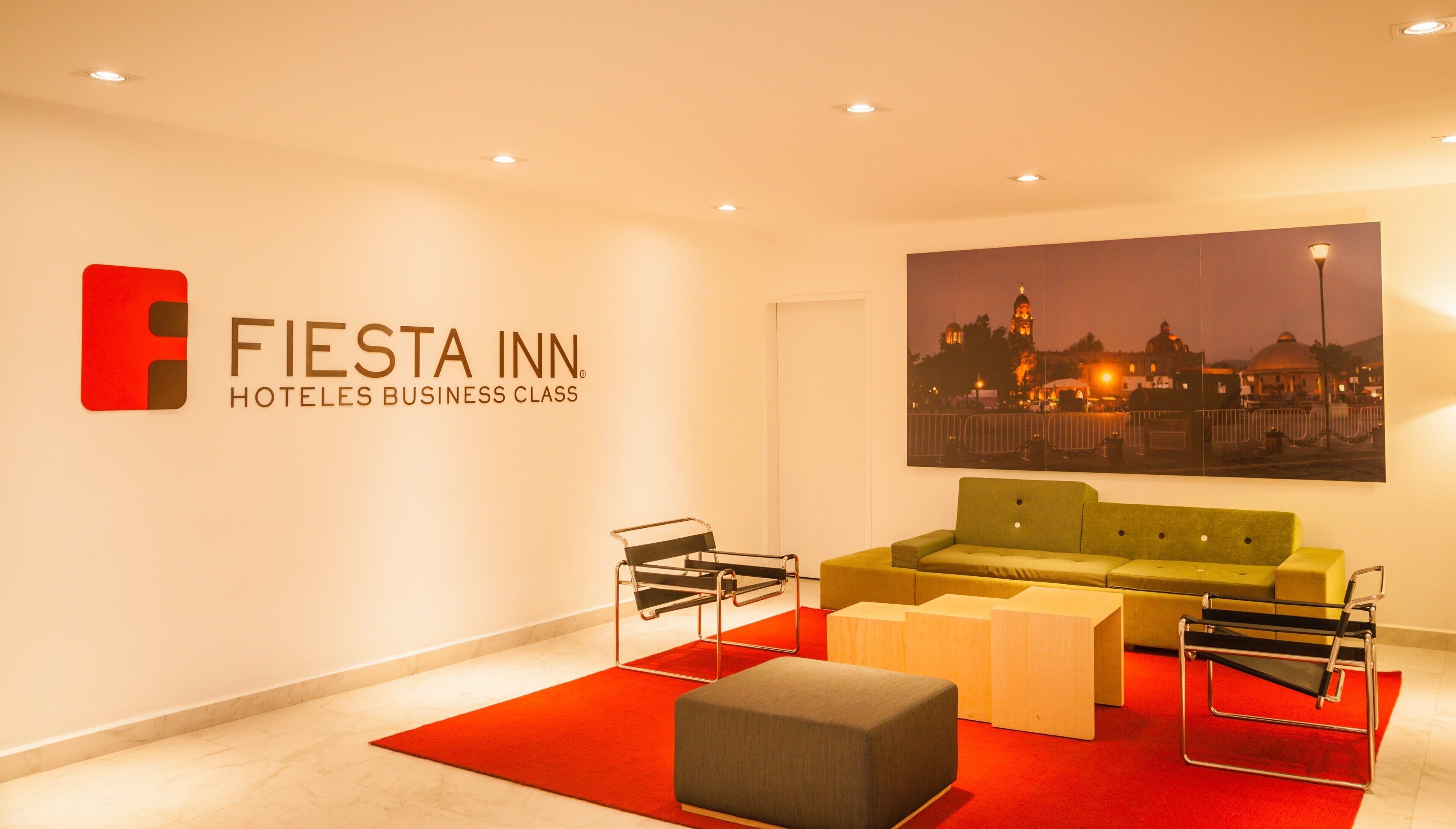 Um saguão de hotel com logo vermelho e branco, um sofá verde, uma cadeira preta e branca, e uma pintura com vista para a cidade.