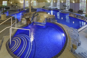 Piscina interna com chão em azulejos azuis, uma formação rochosa e área de estar com cadeiras e mesa.