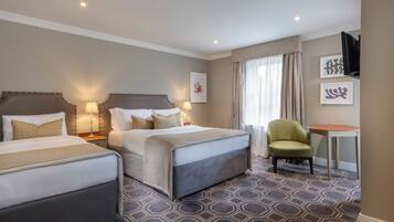Quarto de hotel com duas camas, uma poltrona verde, uma mesa pequena e obras de arte nas paredes.